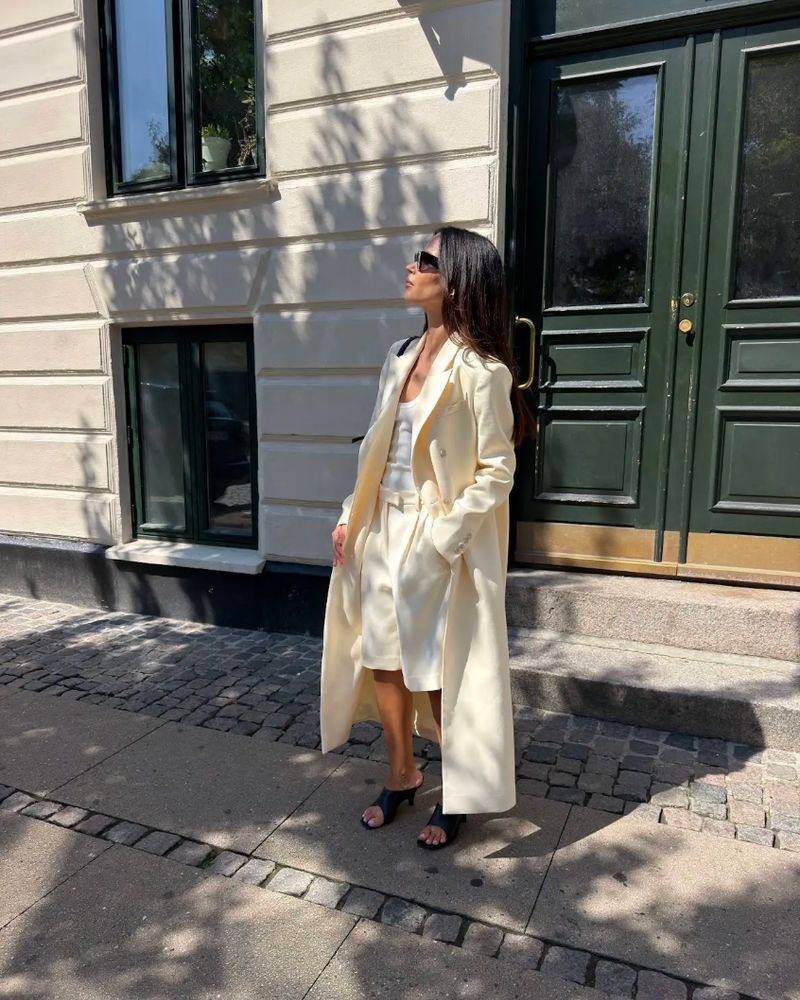 Woman wearing a cream double-breasted long coat with matching tailored shorts and black open-toe heels in a sunny urban setting